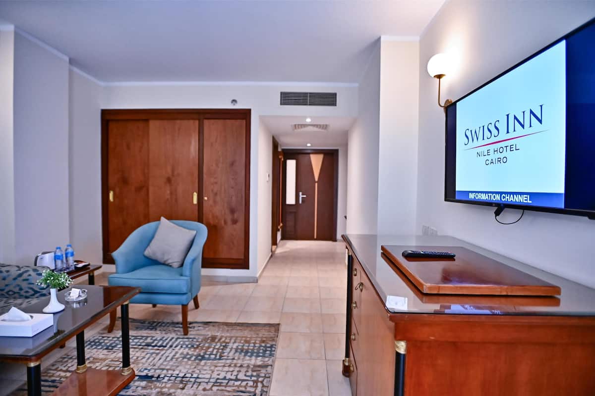 Quarto de hotel com uma televisão grande exibindo o nome do hotel, uma poltrona azul, uma escrivaninha de madeira e um tapete estampado.