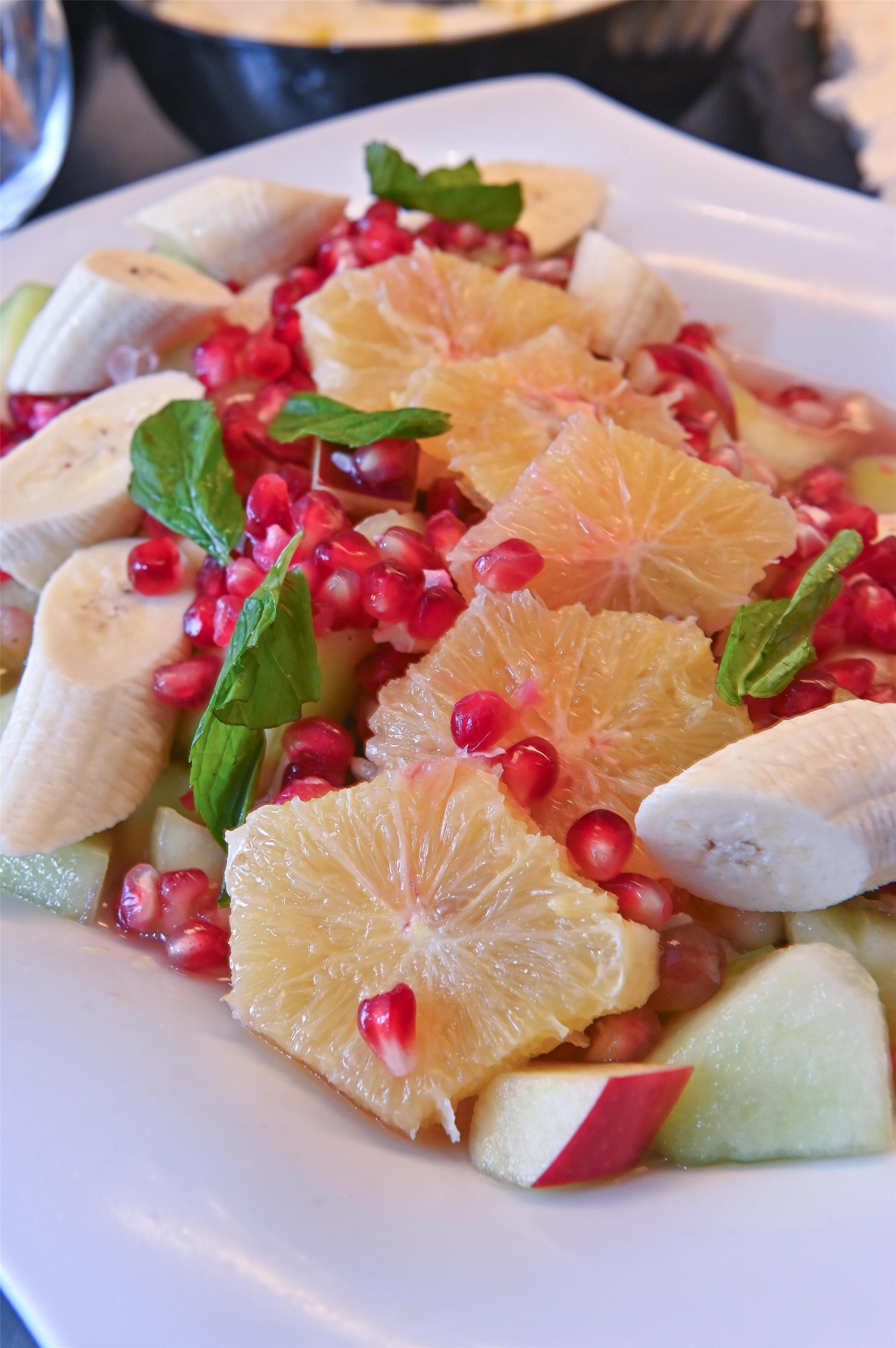 Salada de frutas com bananas, laranjas, sementes de romã e folhas de hortelã em um prato branco.