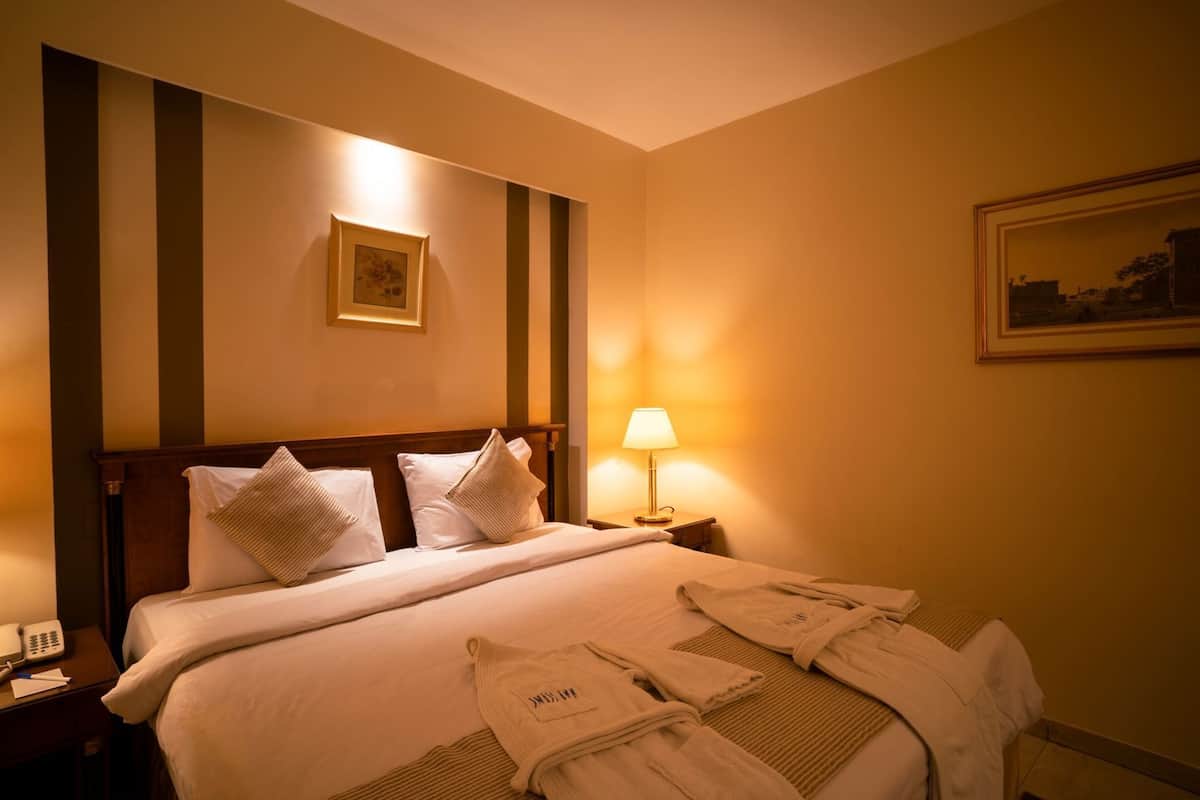 Quarto de hotel com uma cama grande, mesa de cabeceira com abajur e um quadro na parede.