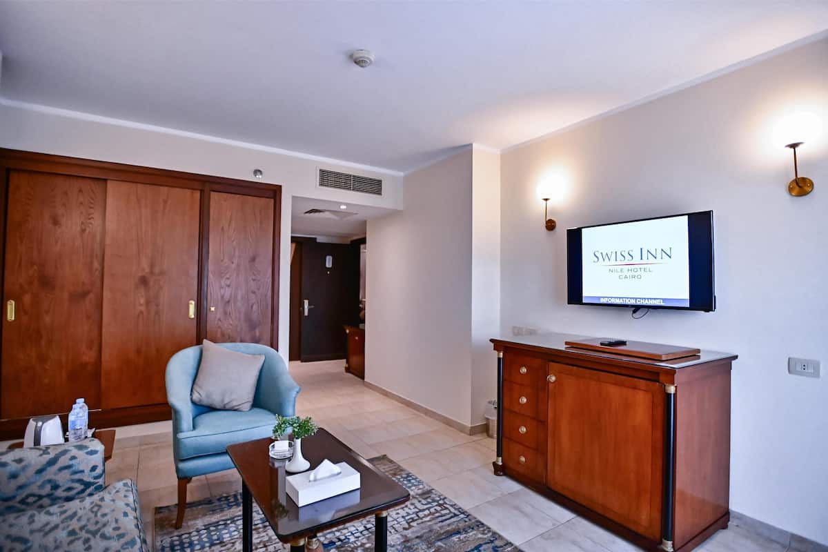 Quarto de hotel com um guarda-roupa de madeira grande, uma poltrona azul, uma mesinha com uma planta e uma televisão na parede exibindo o nome do hotel.