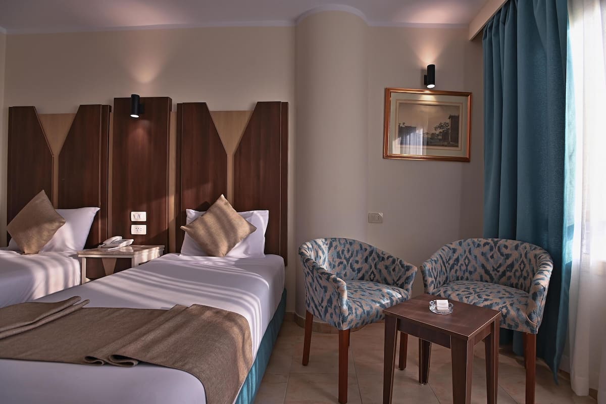 Quarto de hotel com duas camas, uma mesinha, duas cadeiras, uma luminária na parede e um quadro emoldurado.