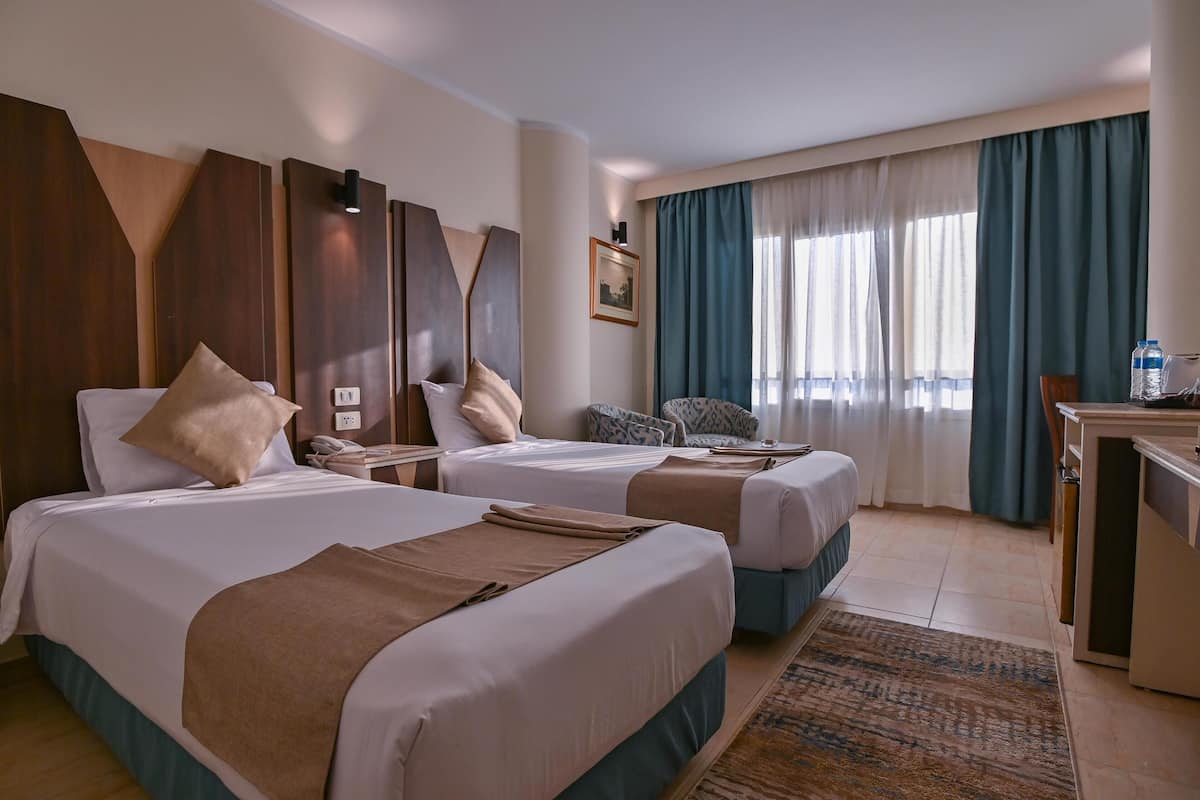 Quarto de hotel com duas camas, uma escrivaninha, uma cadeira e uma janela grande com cortinas.