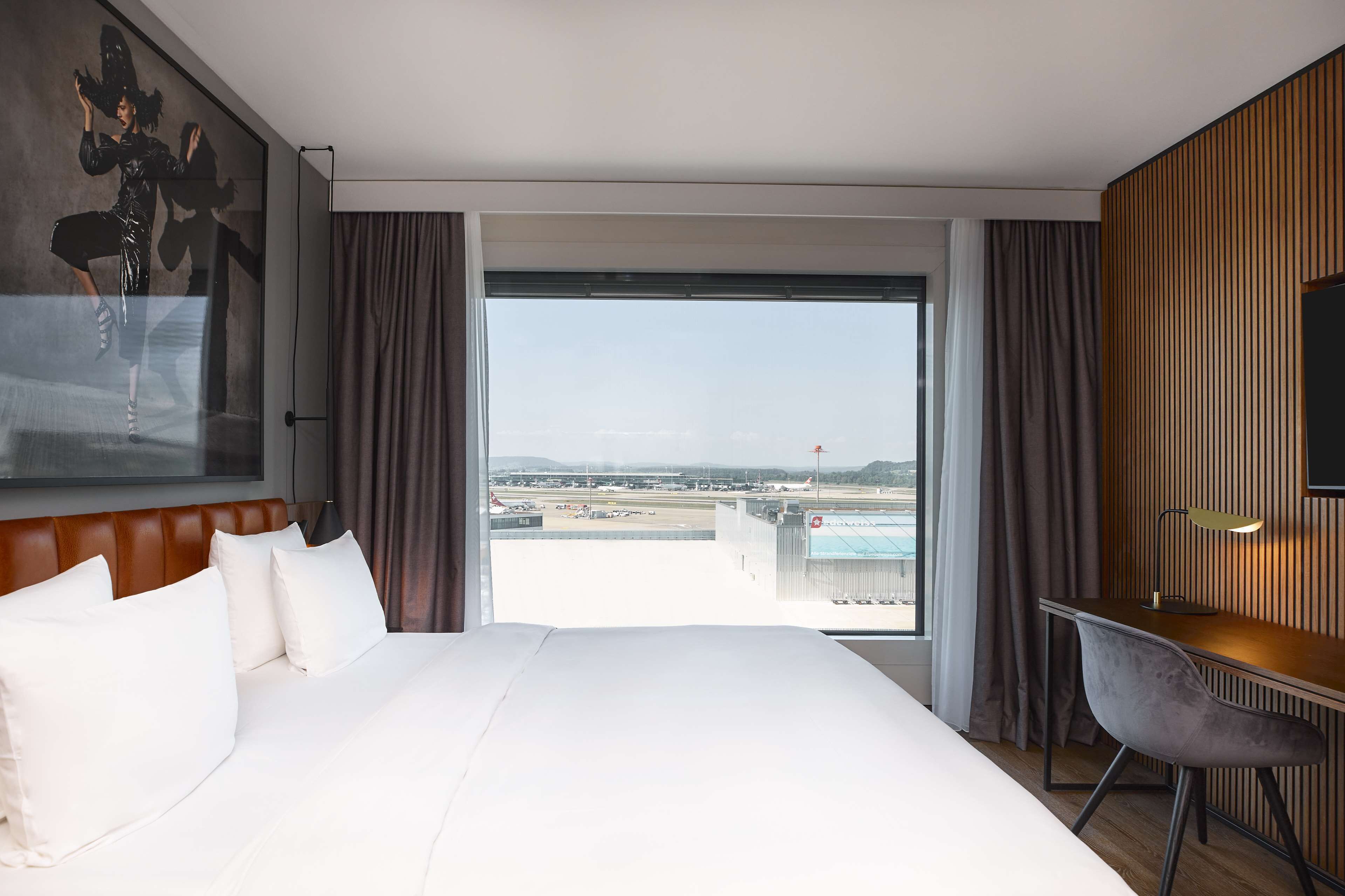 Quarto de hotel com uma cama grande, uma escrivaninha e vista para o aeroporto.