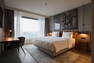 Quarto de hotel moderno com uma cama grande, uma escrivaninha e uma cadeira. Há uma janela ampla com cortinas, um quadro na parede e uma pequena mesa com abajur.