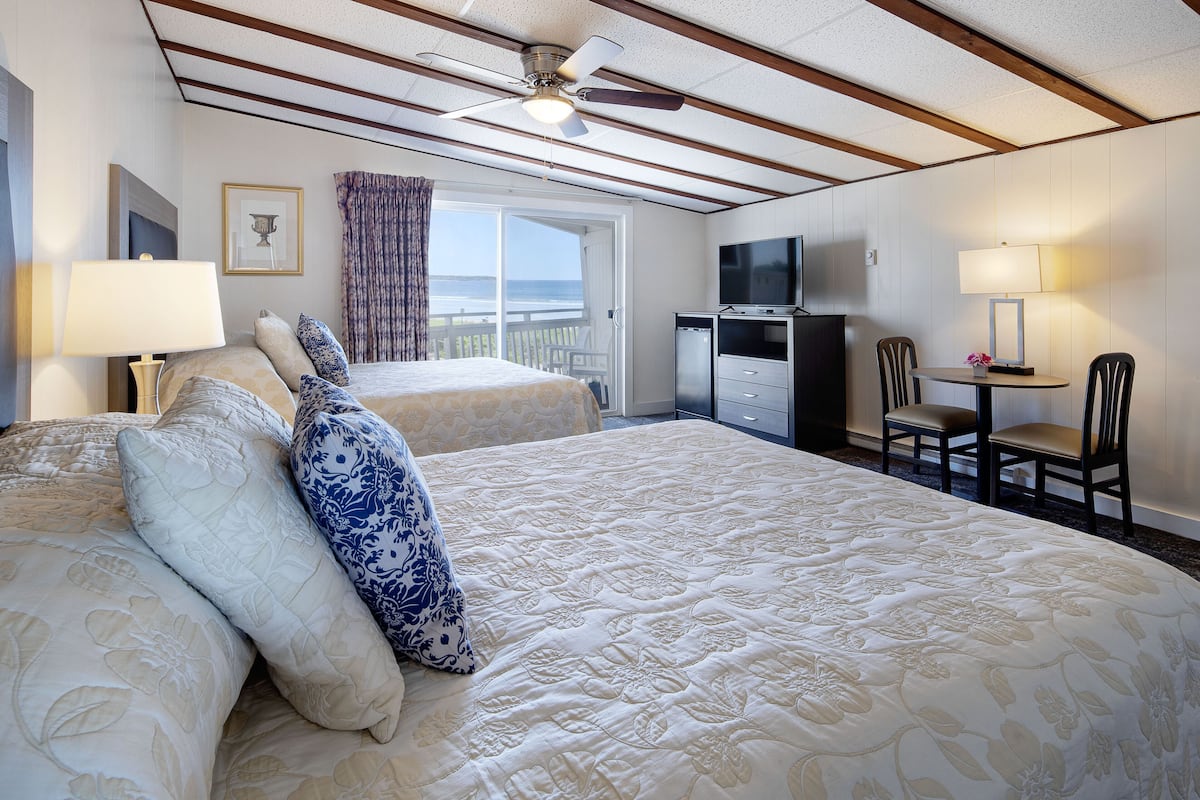 Quarto de hotel com duas camas, ventilador de teto, televisão, escrivaninha com cadeira e vista para o oceano através de uma porta de correr.