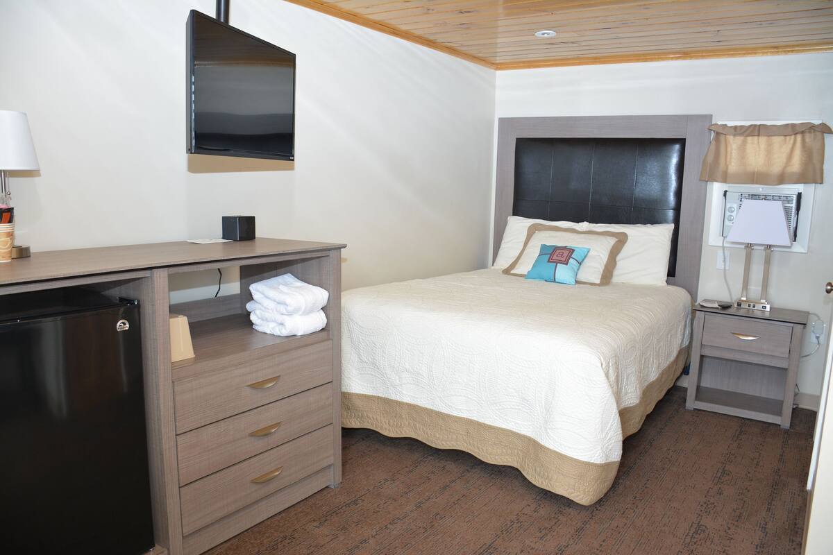 Quarto de hotel com cama, escrivaninha com gavetas, televisão e mesa de cabeceira com abajur.