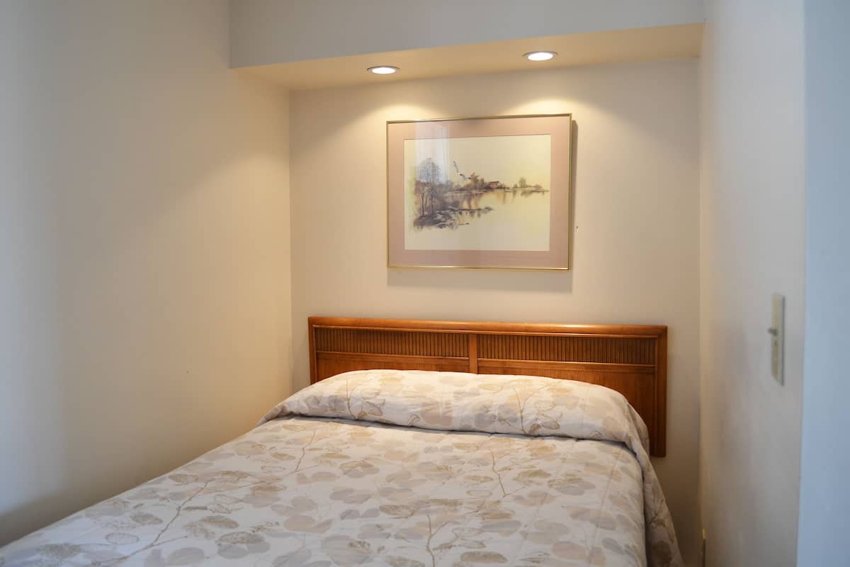 Quarto com cama, cabeceira de madeira, quadro na parede e iluminação embutida no teto.