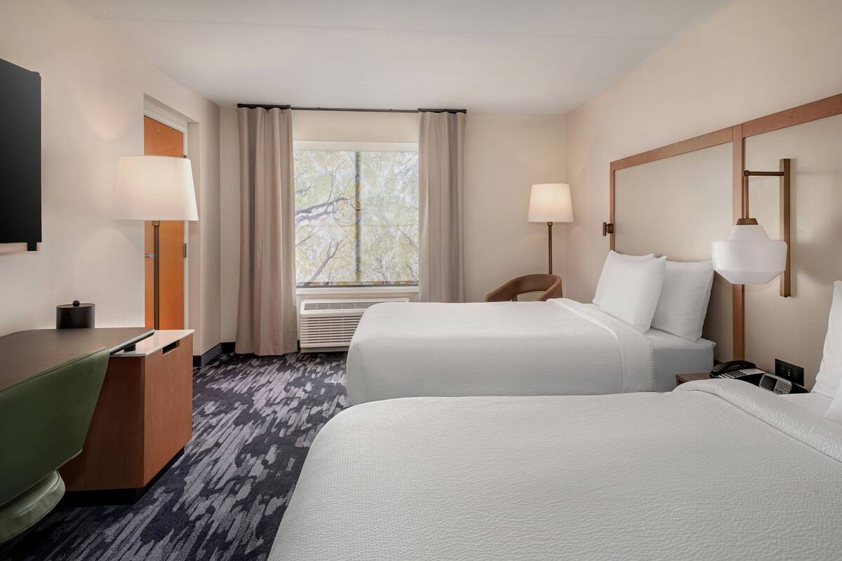 Quarto de hotel com duas camas, uma escrivaninha, uma cadeira e uma janela com cortinas.