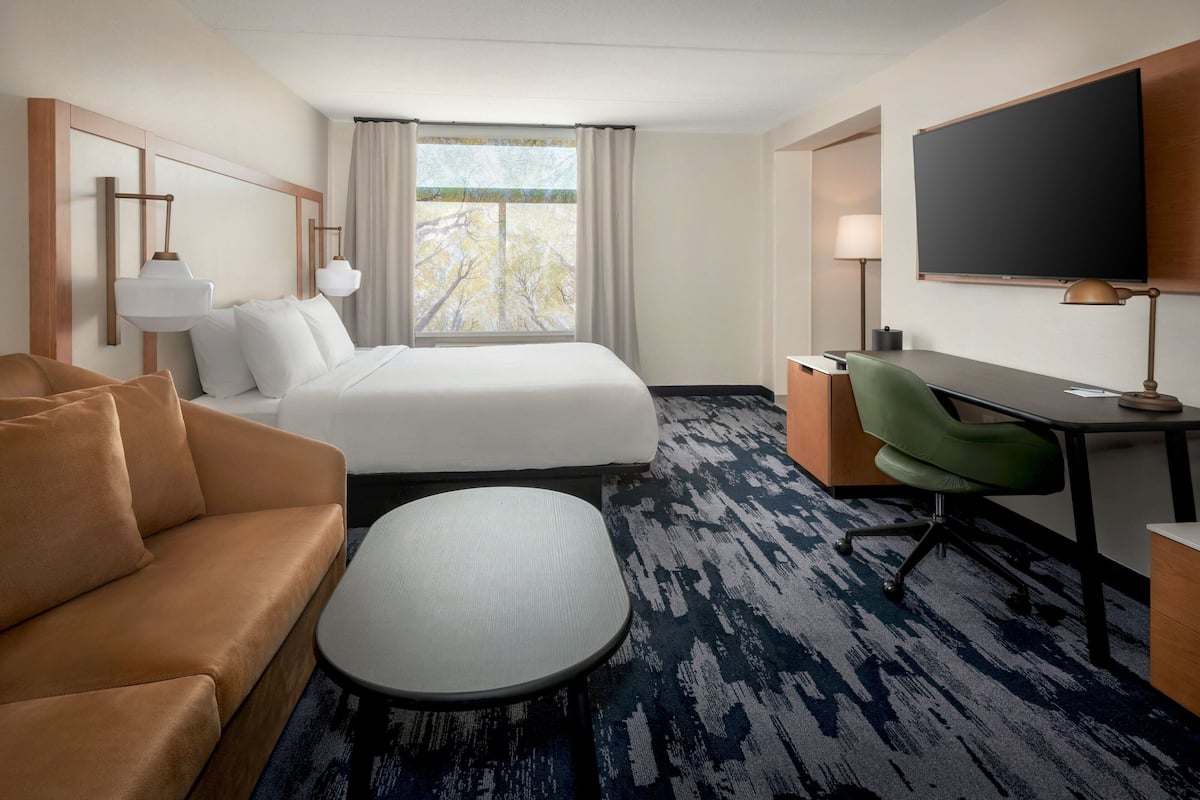 Quarto de hotel com cama, sofá, escrivaninha, cadeira, televisão e janela com cortinas.