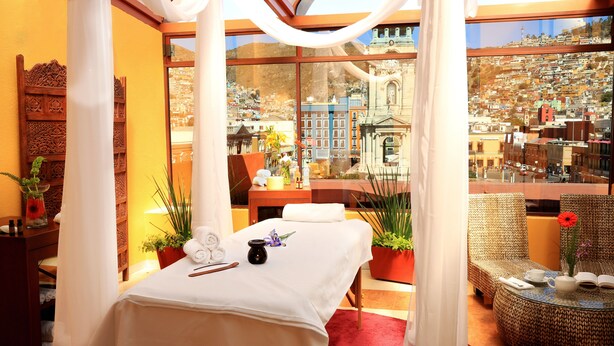 Quarto de hotel com cama, mesa com uma garrafa, uma vela e um vaso com flores. Vê-se uma vista urbana através de amplas janelas.