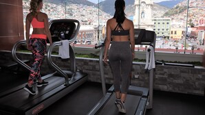 Duas pessoas caminham em esteiras dentro de uma academia, com vista para a cidade e montanhas ao fundo.