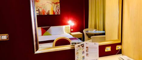 Quarto de hotel com um espelho grande, uma cama com travesseiro vermelho e verde, mesa de cabeceira com abajur, uma cadeira e uma escrivaninha com telefone e folhetos.