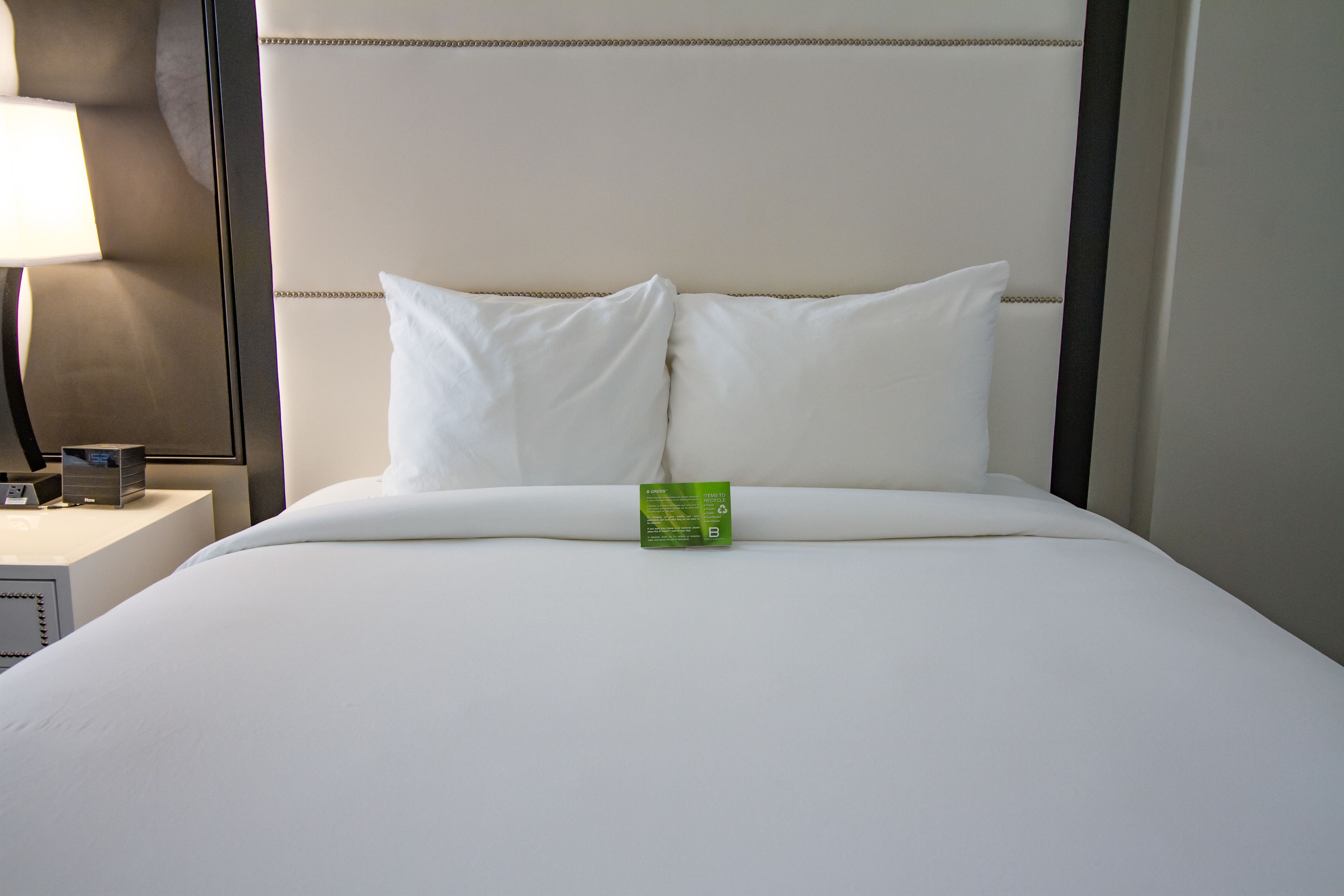 Cama bem arrumada com lençóis e almofadas brancos, mesa de cabeceira com abajur e uma placa verde sobre a cama.