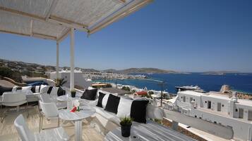 Terraço com mobiliário branco, vista para o mar e uma vila costeira ao fundo.