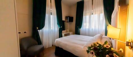 Quarto de hotel com uma cama, escrivaninha, cadeira, televisão e luminária.