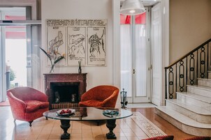 Uma sala de estar com dois poltronas vermelhas, uma mesa de centro de vidro e uma lareira. Há uma escada com corrimão preto e uma porta com cortina vermelha.