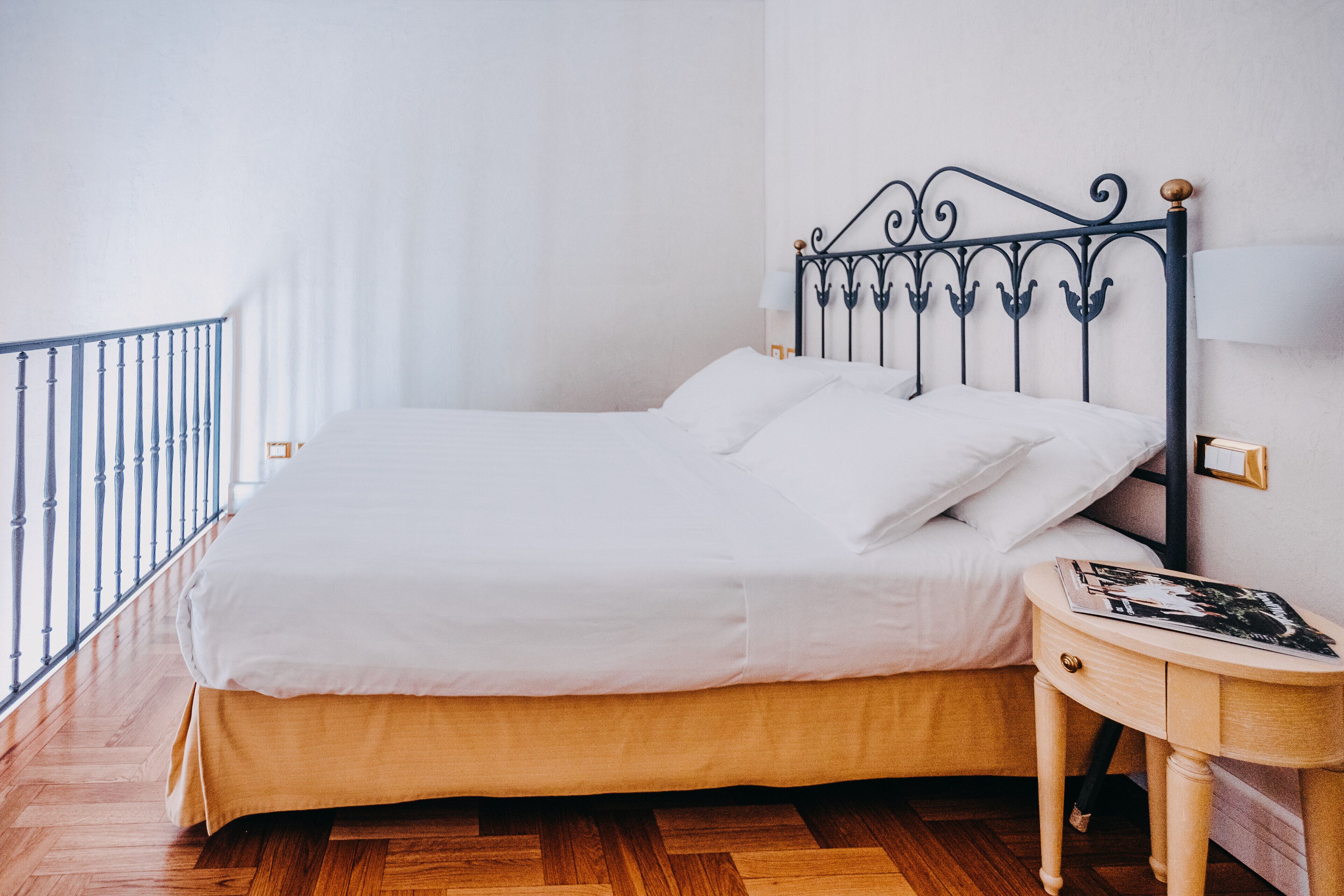 Cama bem arrumada com lençóis brancos, cabeceira de madeira e pequena mesa de cabeceira com abajur.