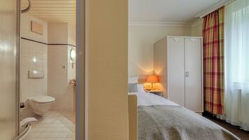 Casa de banho com sanita, lavatório e espelho. O quarto tem uma cama com um cobertor e uma mesa de cabeceira com uma lâmpada. Há um guarda-roupa e uma janela com cortinas.