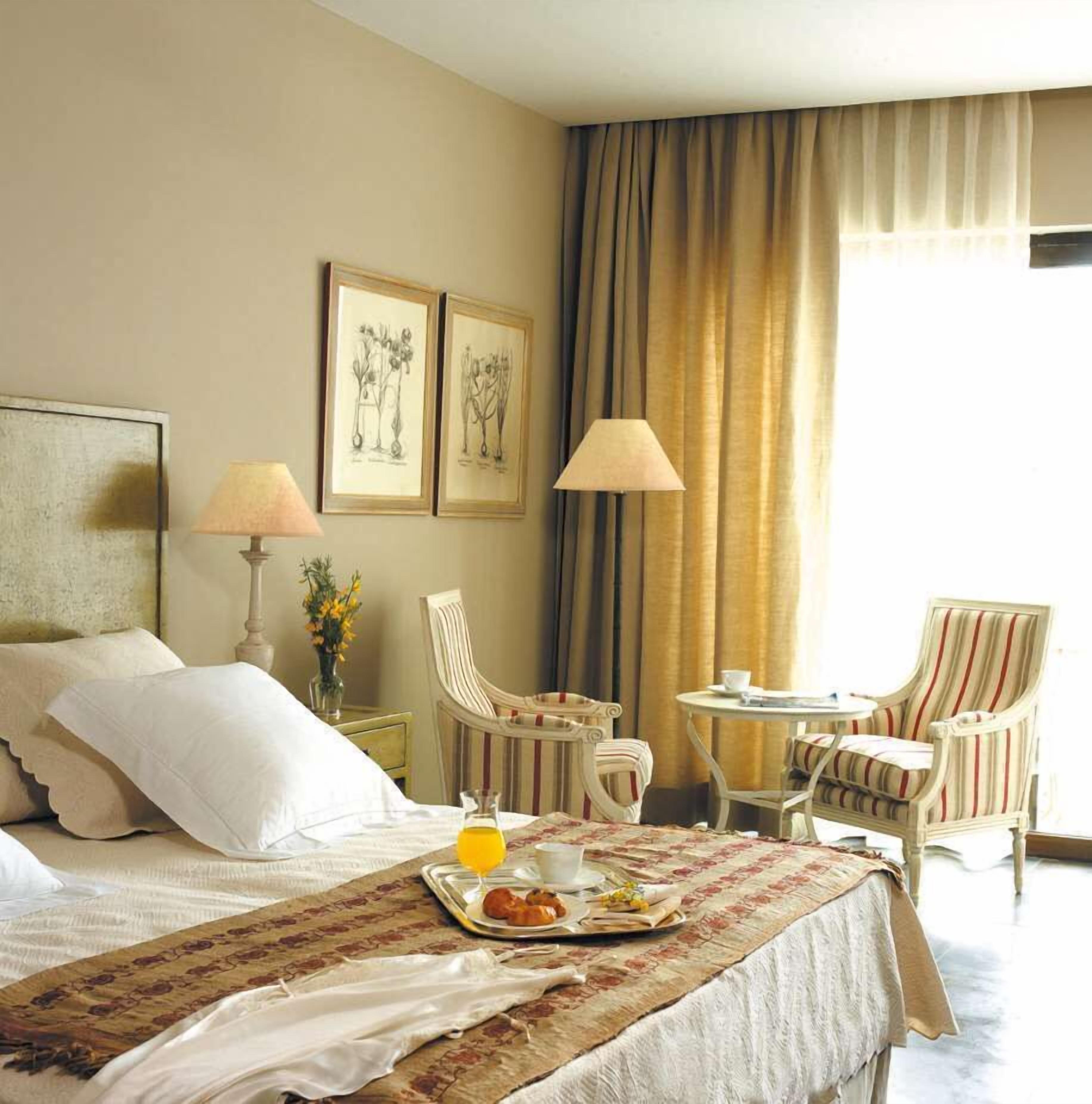 Quarto de hotel com cama, duas cadeiras, uma mesinha e uma bandeja com café da manhã.