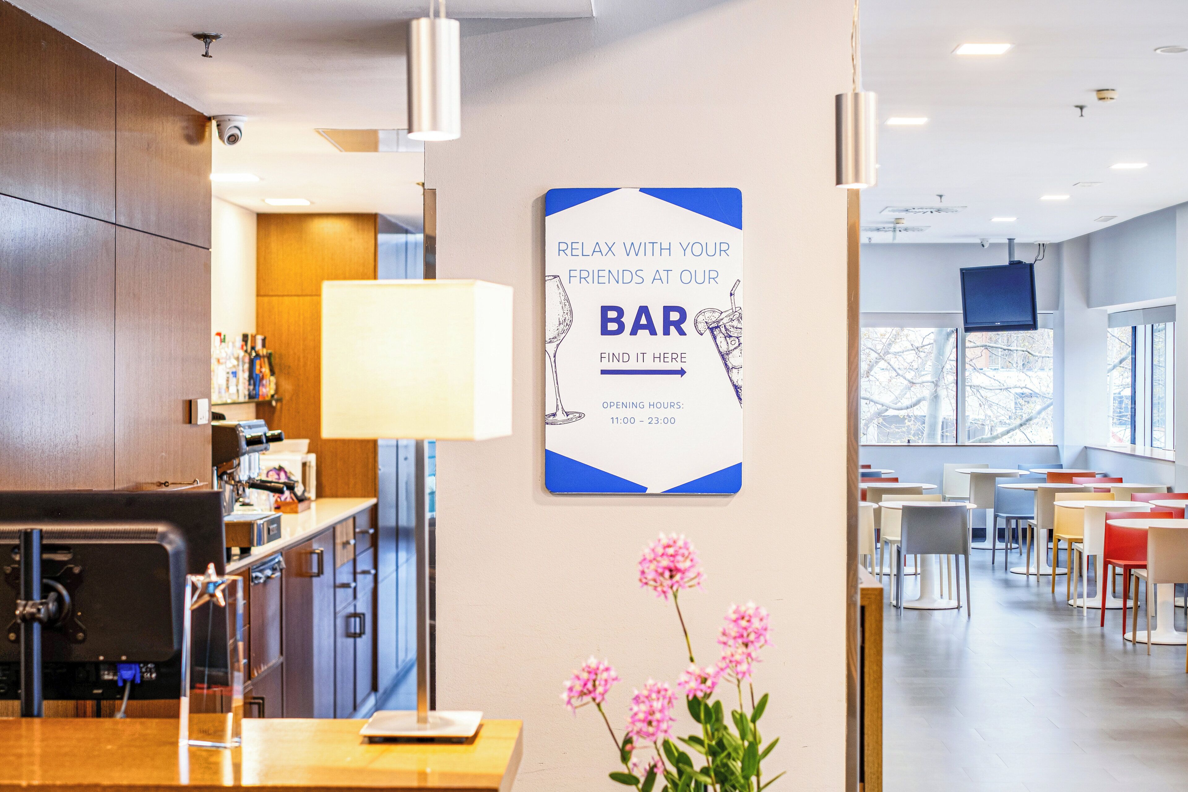 Uma área de bar com um aviso indicando ser para relaxar com amigos, situada em um hall de hotel moderno com um balcão de recepção de madeira e vista para o exterior.