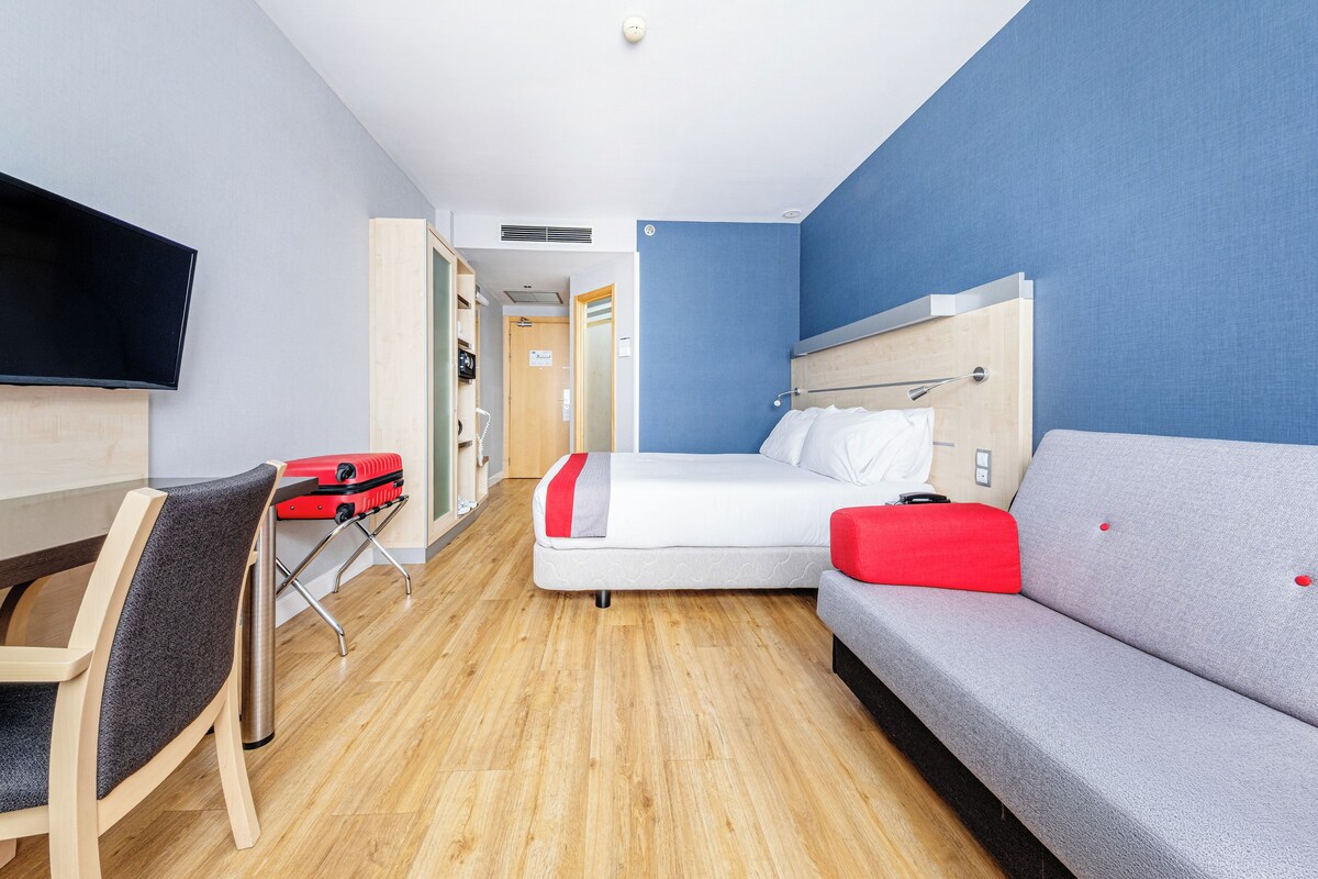 Quarto de hotel com cama, sofá, escrivaninha e televisão.
