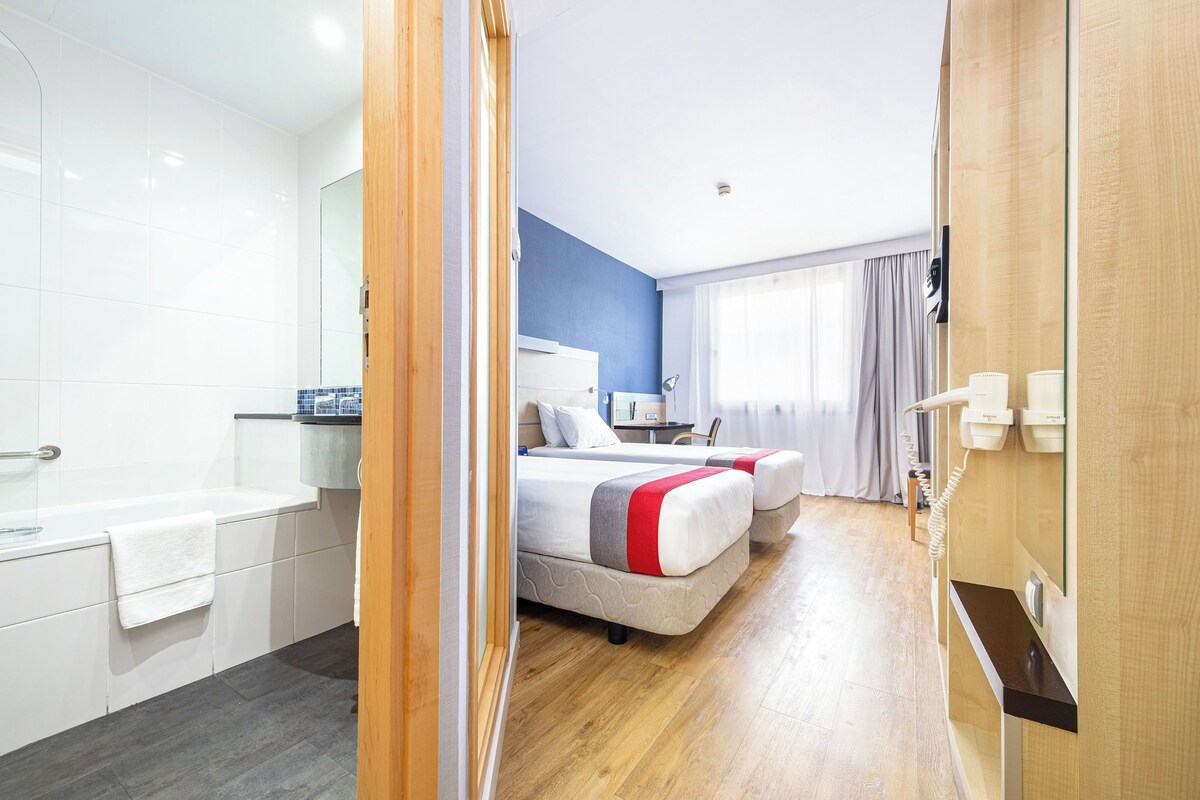Quarto de hotel moderno com banheira, cama de casal, escrivaninha e uma grande janela com cortinas.