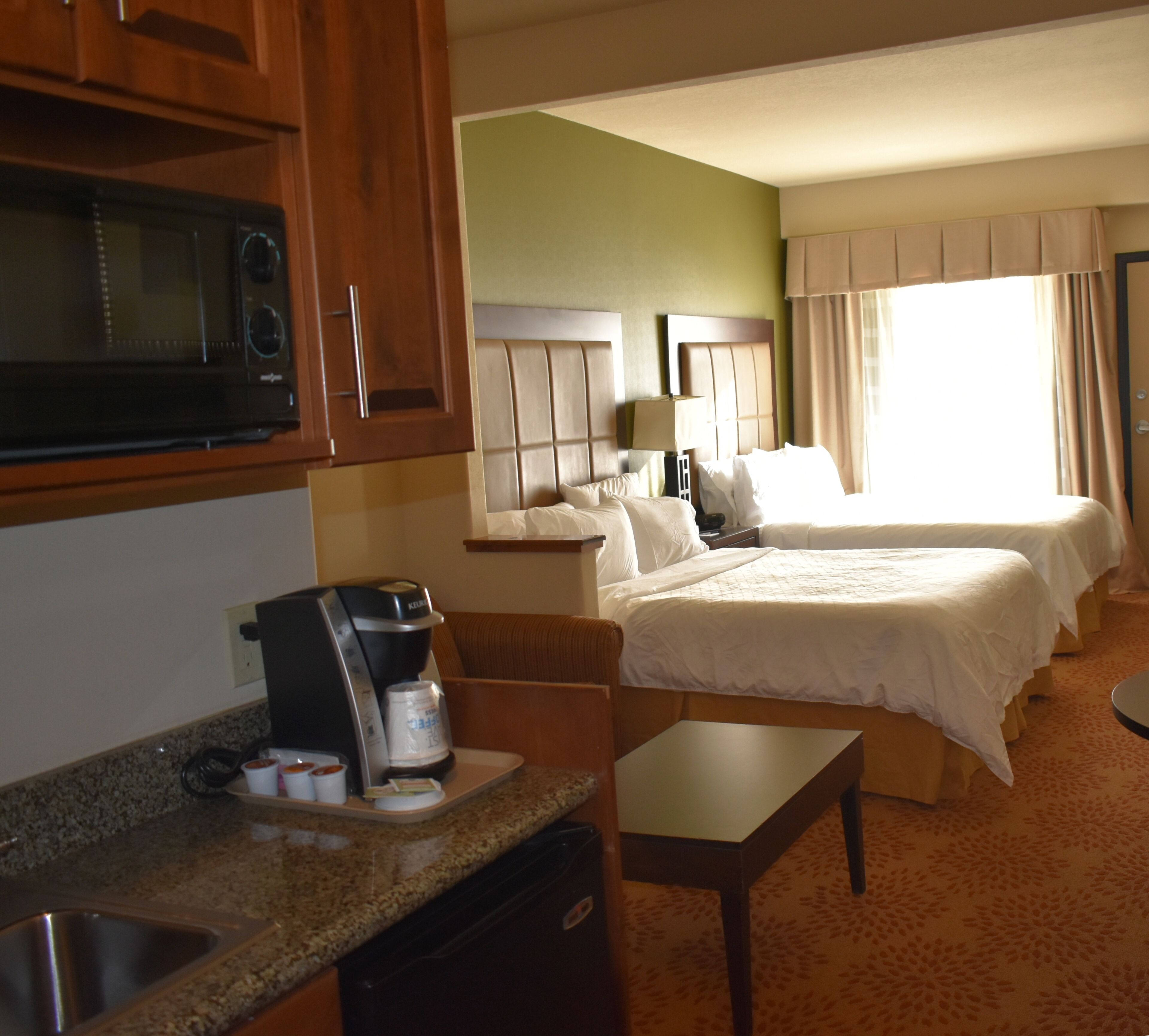 Quarto de hotel com cozinha compacta, uma cama, mesa de cabeceira e janela com cortinas.