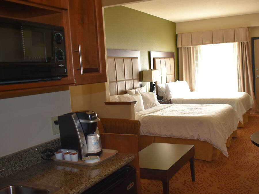 Quarto de hotel com cozinha compacta, uma cama, mesa de cabeceira e janela com cortinas.