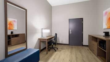 Quarto de hotel com uma escrivaninha de madeira, uma cadeira, um banco azul, um micro-ondas, uma máquina de café e uma porta.
