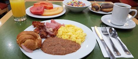 Disposição de café da manhã com um prato de ovos, bacon, presunto e um croissant, uma tigela de cereal, um copo de suco de laranja e uma xícara de café sobre uma mesa.