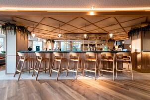 Uma área de bar moderna com balcão de madeira, vários bancos altos, luminárias pendentes e prateleiras com copos.