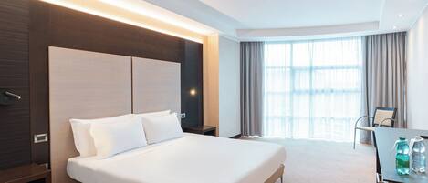 Quarto de hotel moderno com uma cama grande, uma escrivaninha com cadeira e um janelão com cortinas.