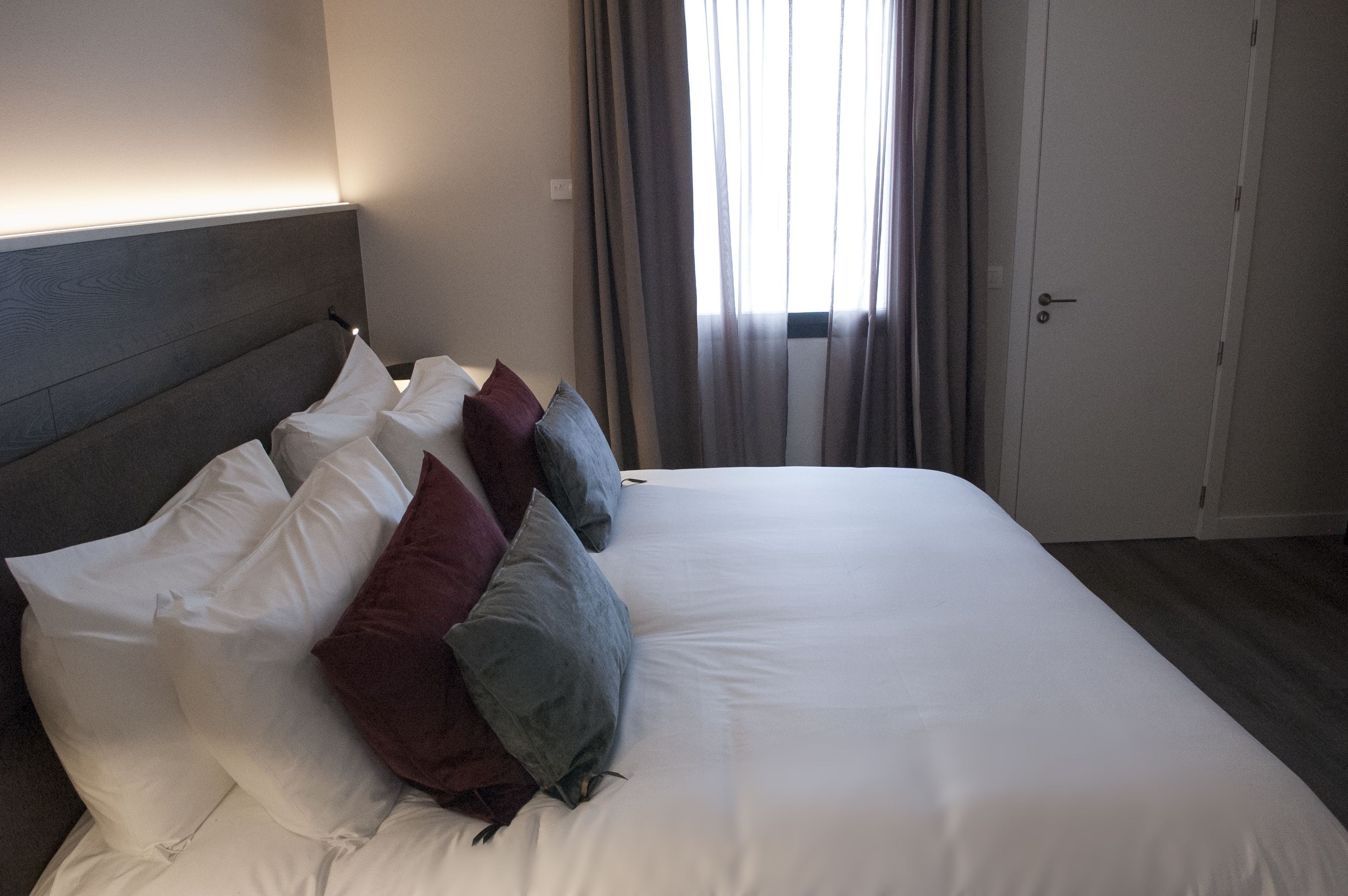 Cama bem arrumada com lençóis brancos e cinzentos, cabeceira cinza e vários travesseiros em cores variadas.