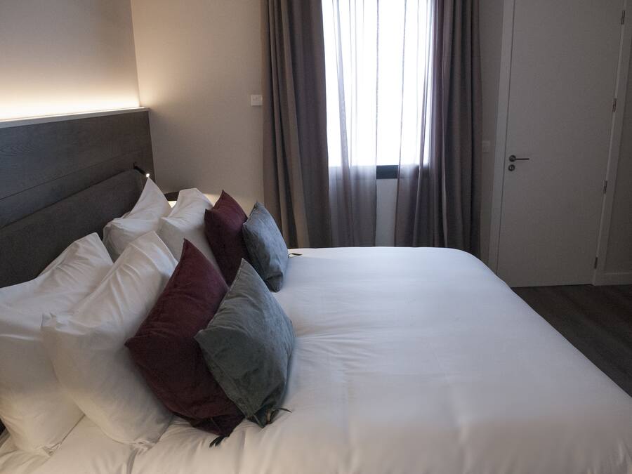 Cama bem arrumada com lençóis brancos e cinzentos, cabeceira cinza e vários travesseiros em cores variadas.