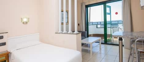 Quarto de hotel com cama, uma mesinha, uma cadeira, varanda com mesa e cadeiras, e janela com cortinas.