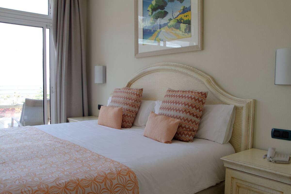 Quarto de hotel com uma cama grande, duas almofadas estampadas, mesa de cabeceira com telefone e um quadro na parede.
