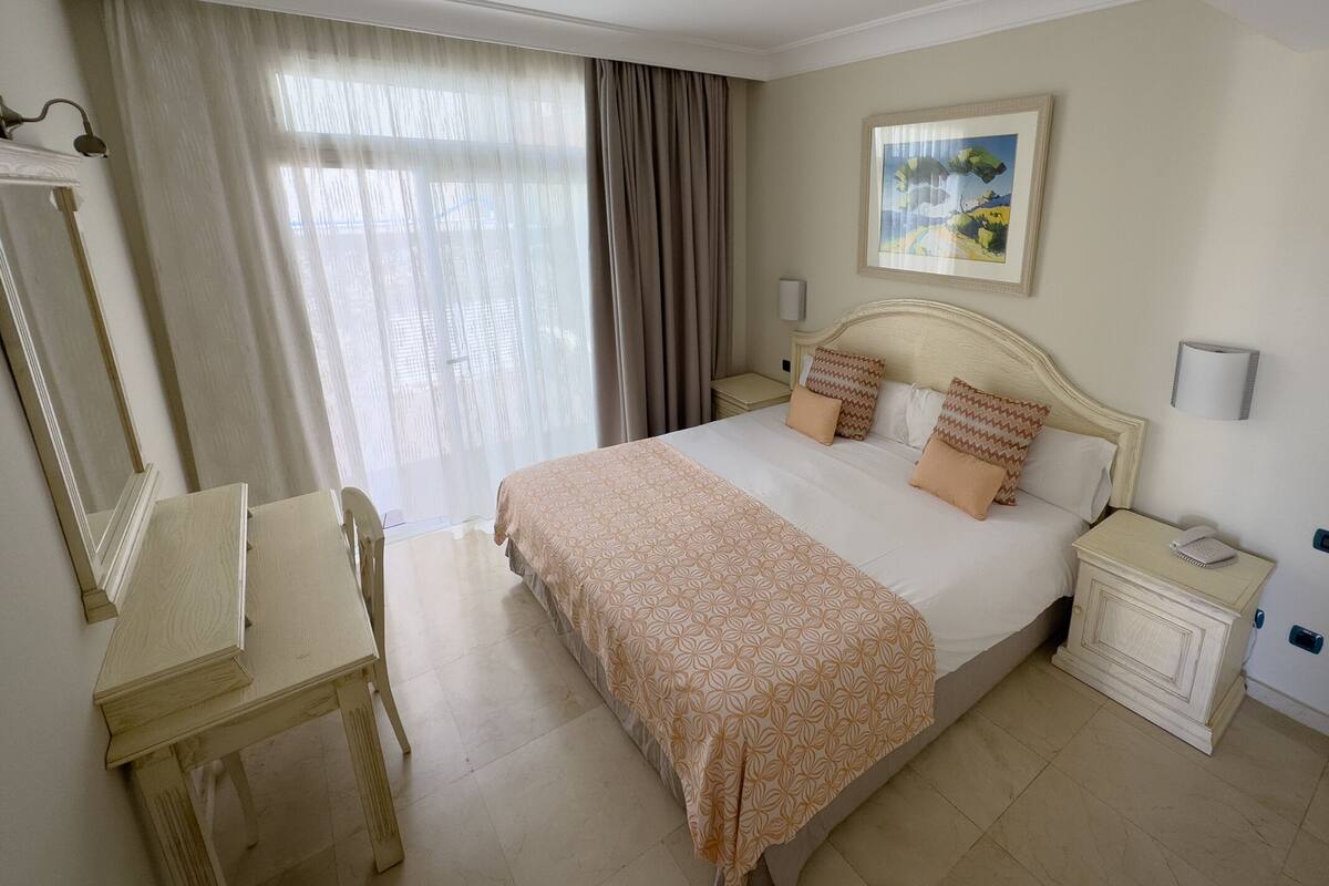 Quarto de hotel com cama, mesas de cabeceira, escrivaninha, cadeira e um quadro na parede.