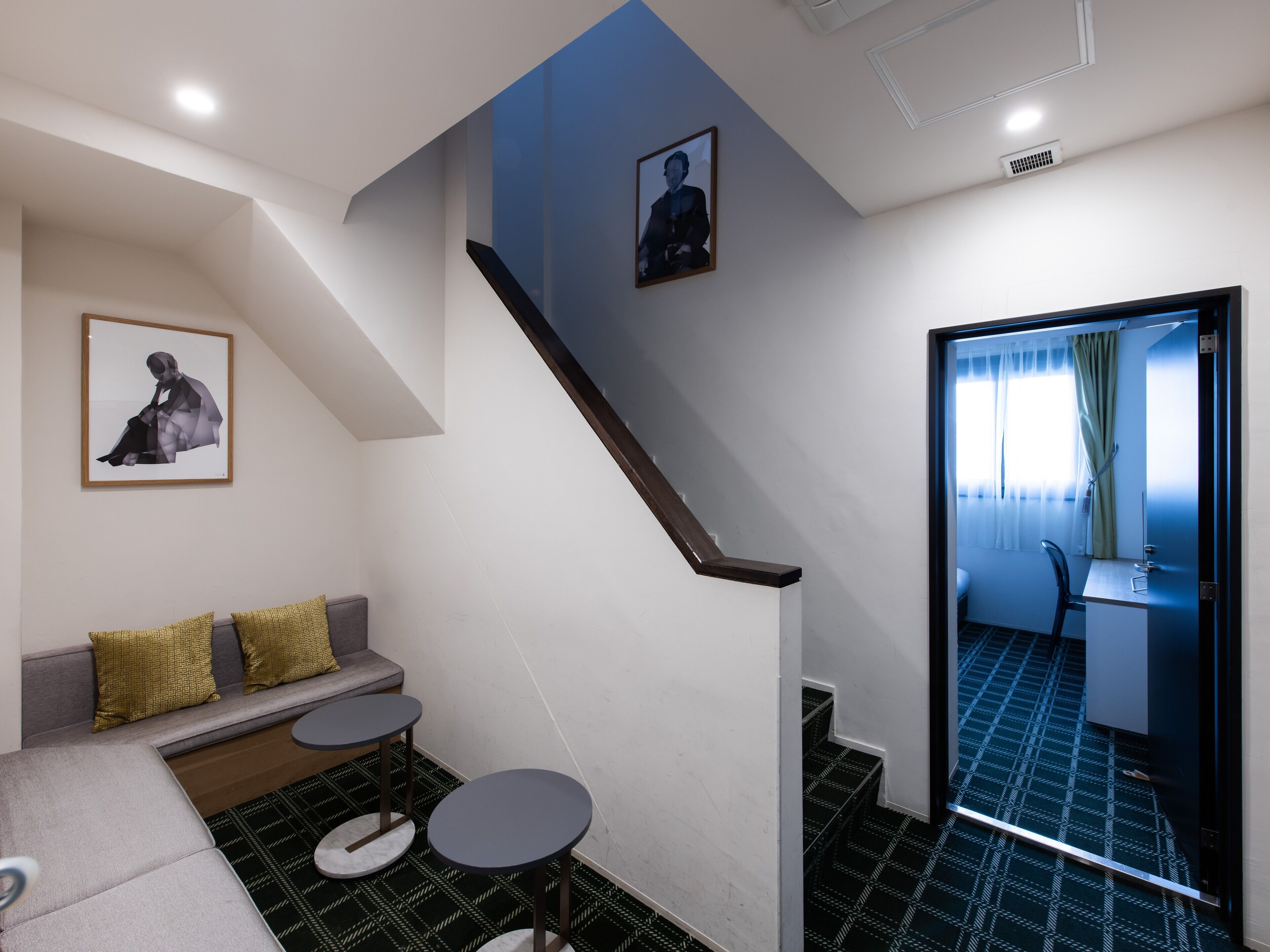 Quarto de hotel moderno com escada, sofá com almofadas, duas mesinhas e um quadro emoldurado na parede.