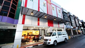 Uma carrinha branca estacionada em frente a um edifício com fachada vermelha e branca, uma área de estar ao ar livre coberta e uma placa indicando um restaurante ou café.