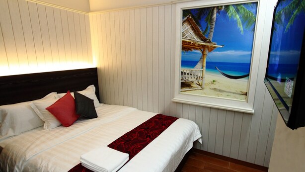 Cama bem arrumada com travesseiro vermelho e preto, colcha branca e vermelha, e janela com pintura de uma paisagem praiana.