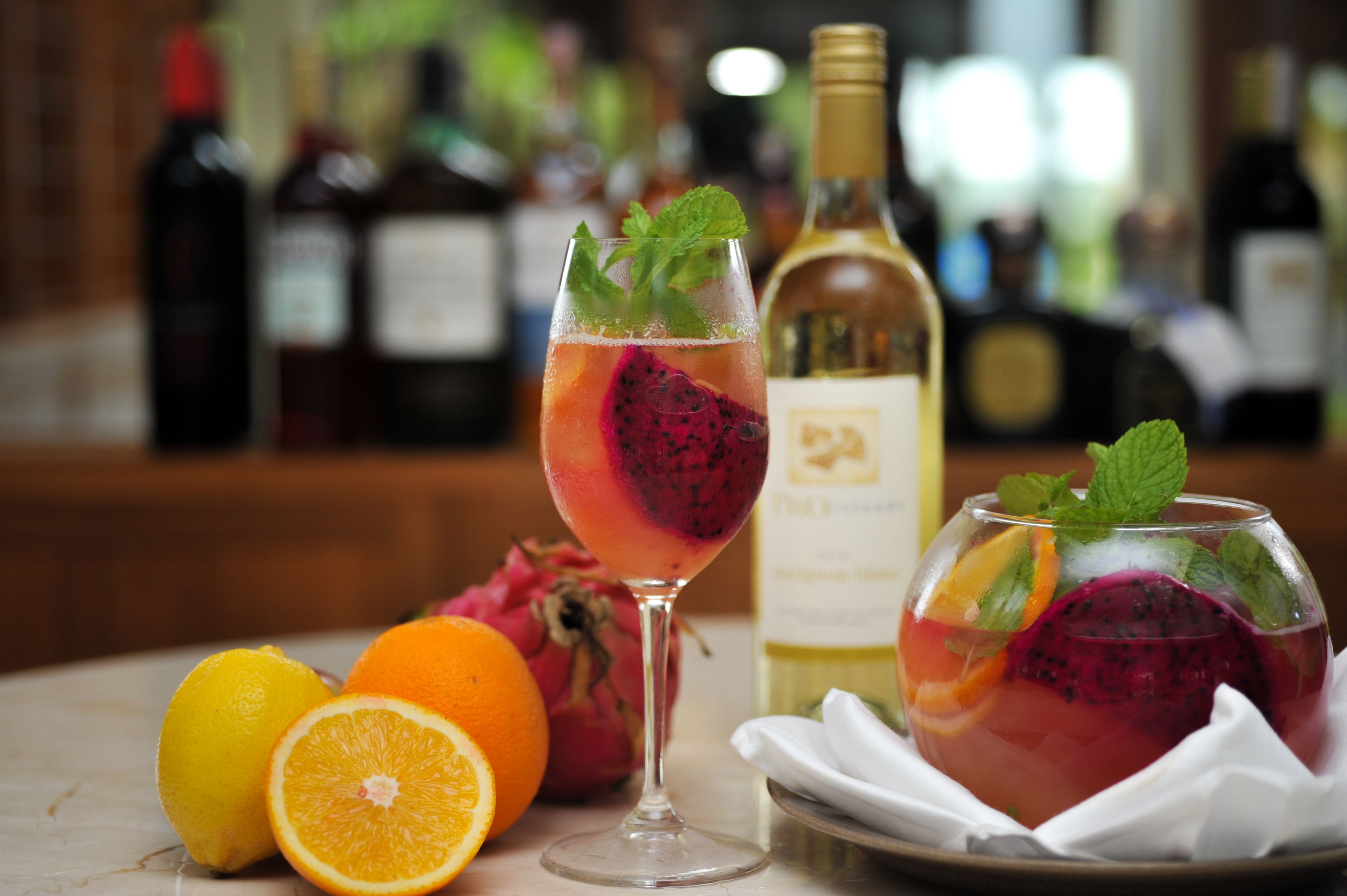 Um bar com um copo de cocktail decorado com frutas e hortelã, uma garrafa de vinho e laranjas frescas sobre a mesa.
