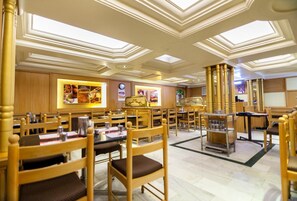Restaurante com cadeiras e mesas de madeira, área de bar central e padrões decorativos no teto.