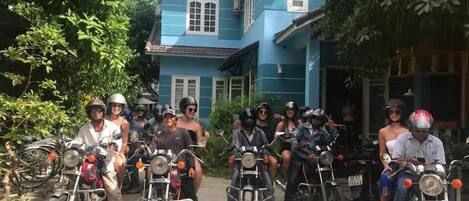 Um grupo de pessoas em motocicletas estacionadas em frente a uma casa azul com telhado de azulejos.