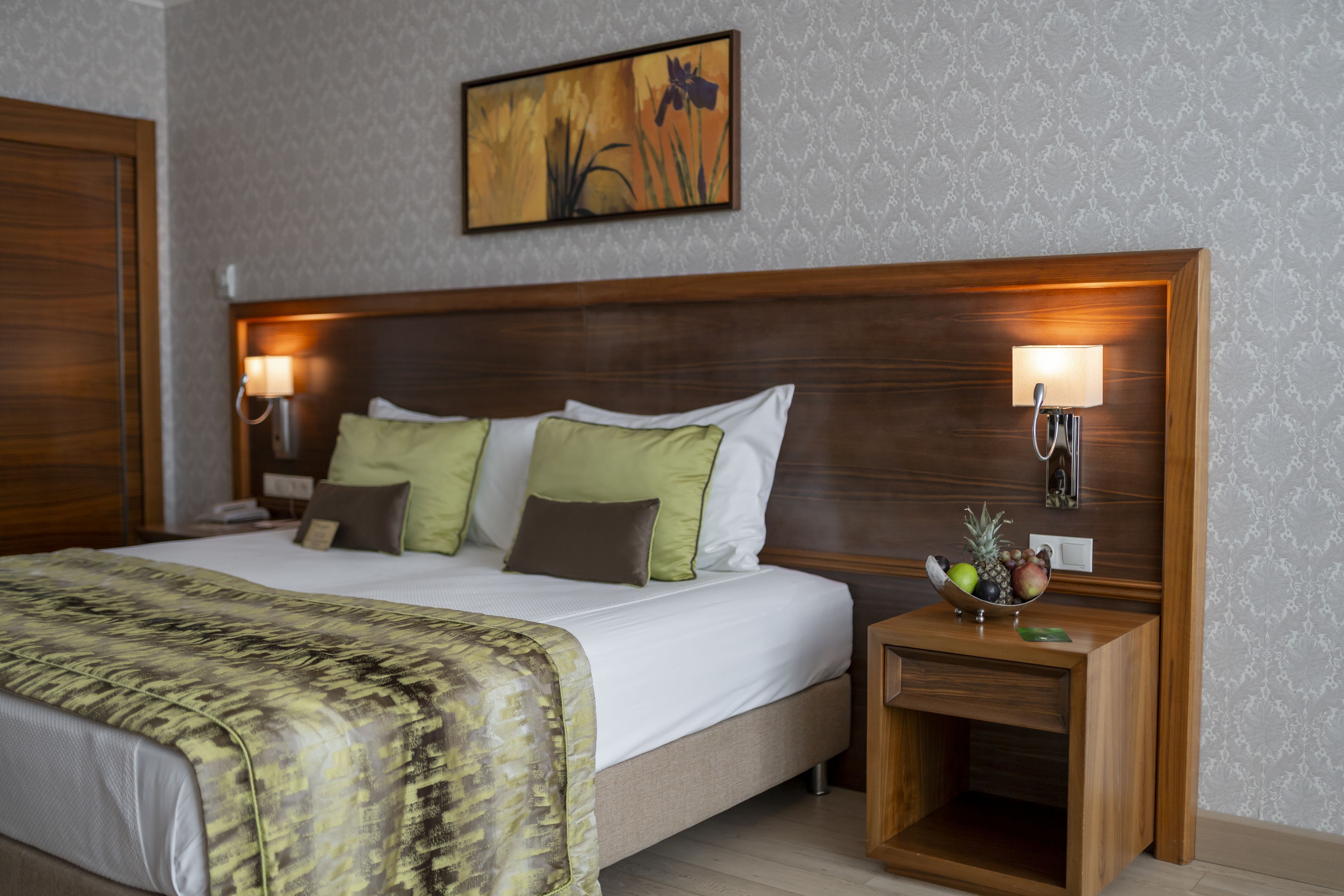 Quarto de hotel com uma cama grande, duas luminárias de cabeceira, uma mesinha de cabeceira com uma tigela de frutas e uma obra de arte emoldurada na parede.