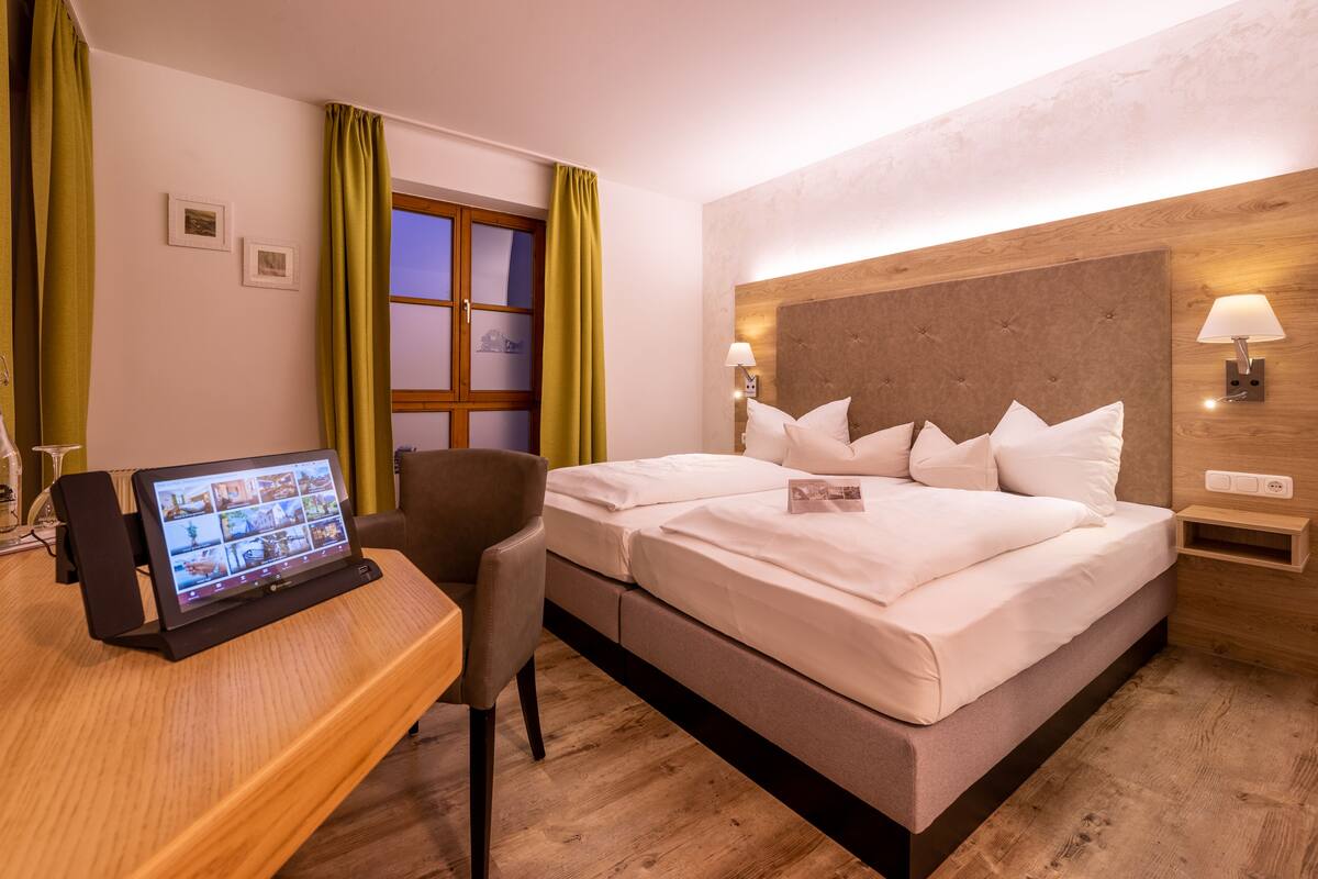 Quarto de hotel com cama, escrivaninha com monitor de computador, cadeira e janela com cortinas.