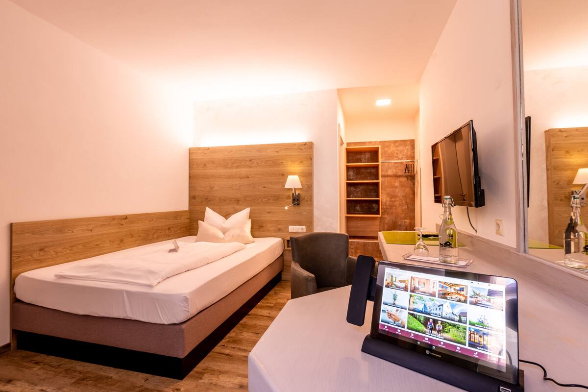 Quarto de hotel com cama de cabeceira de madeira, escrivaninha com um laptop, cadeira, televisão e estante com garrafas.