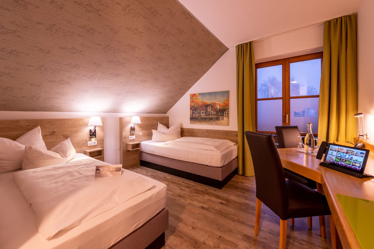 Quarto de hotel com duas camas, uma mesa de trabalho com um laptop, um quadro na parede e uma janela com cortinas.