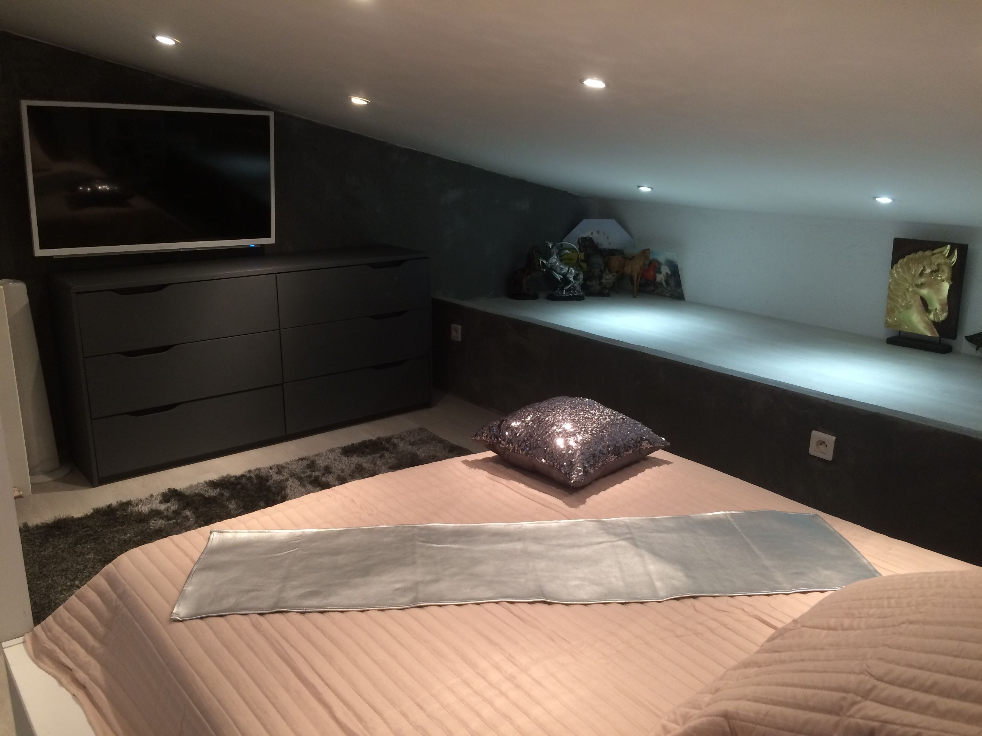 Quarto com cama, penteadeira, televisão e prateleira com objetos decorativos.