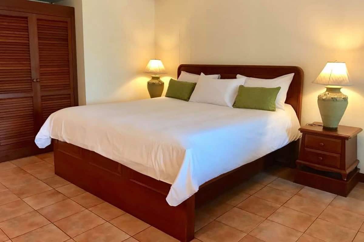 Cama bem arrumada com lençóis brancos e almofadas verdes, com duas mesas de cabeceira com luminárias de cada lado.