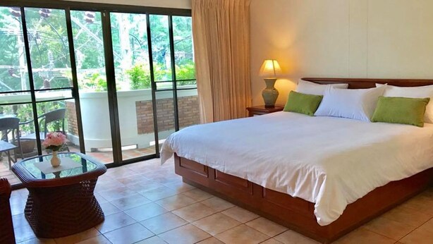 Quarto de hotel com uma cama grande, uma varanda com mesa e cadeiras, e vista para o verde lá fora.